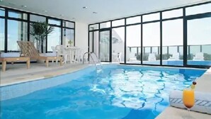 Piscina interior moderna com paredes de vidro transparente, cadeiras de descanso e um copo de sumo de laranja junto à piscina.