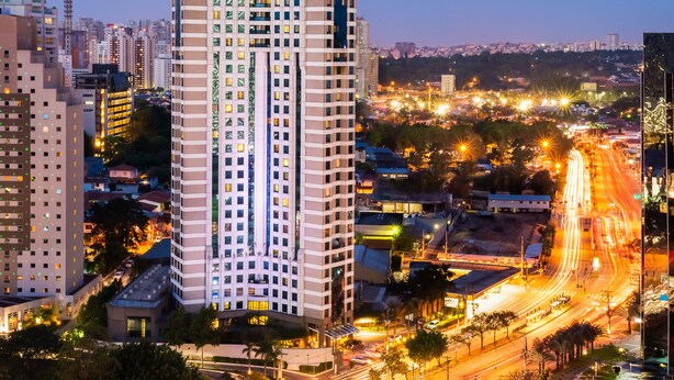 Um edifício alto com design moderno, cercado por uma movimentada rua urbana com tráfego visível.