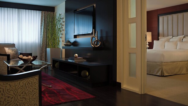 Quarto de hotel moderno com piso de madeira escura, tapete vermelho, mesa de centro de vidro, sofá, TV fixada na parede e cama com roupa de cama branca.