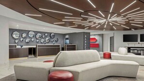 Um lobby moderno com sofá em seção, pufes vermelhos, balcão de recepção e TV fixada na parede.