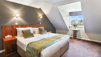 Quarto moderno com uma cama grande, cabeceira de madeira e mesa de cabeceira. Há uma pequena mesa com um vaso de flores e uma janela com vista para vegetação.