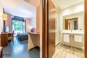 Quarto de hotel com escrivaninha, cadeira, cama e banheiro com pia e espelho.