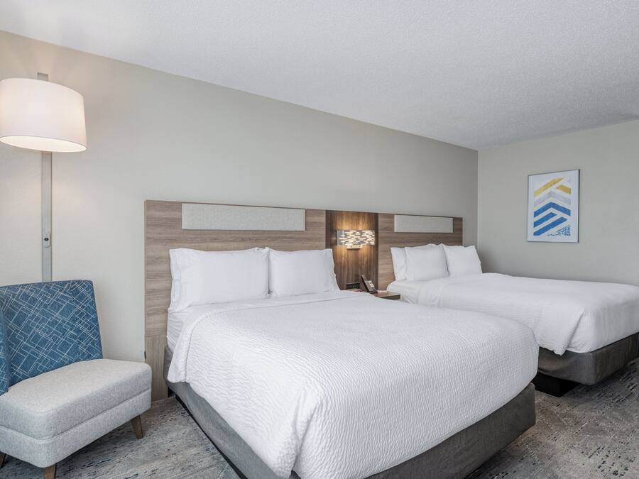 Quarto de hotel com duas camas, uma poltrona azul, mesa de cabeceira com abajur e uma obra de arte na parede.