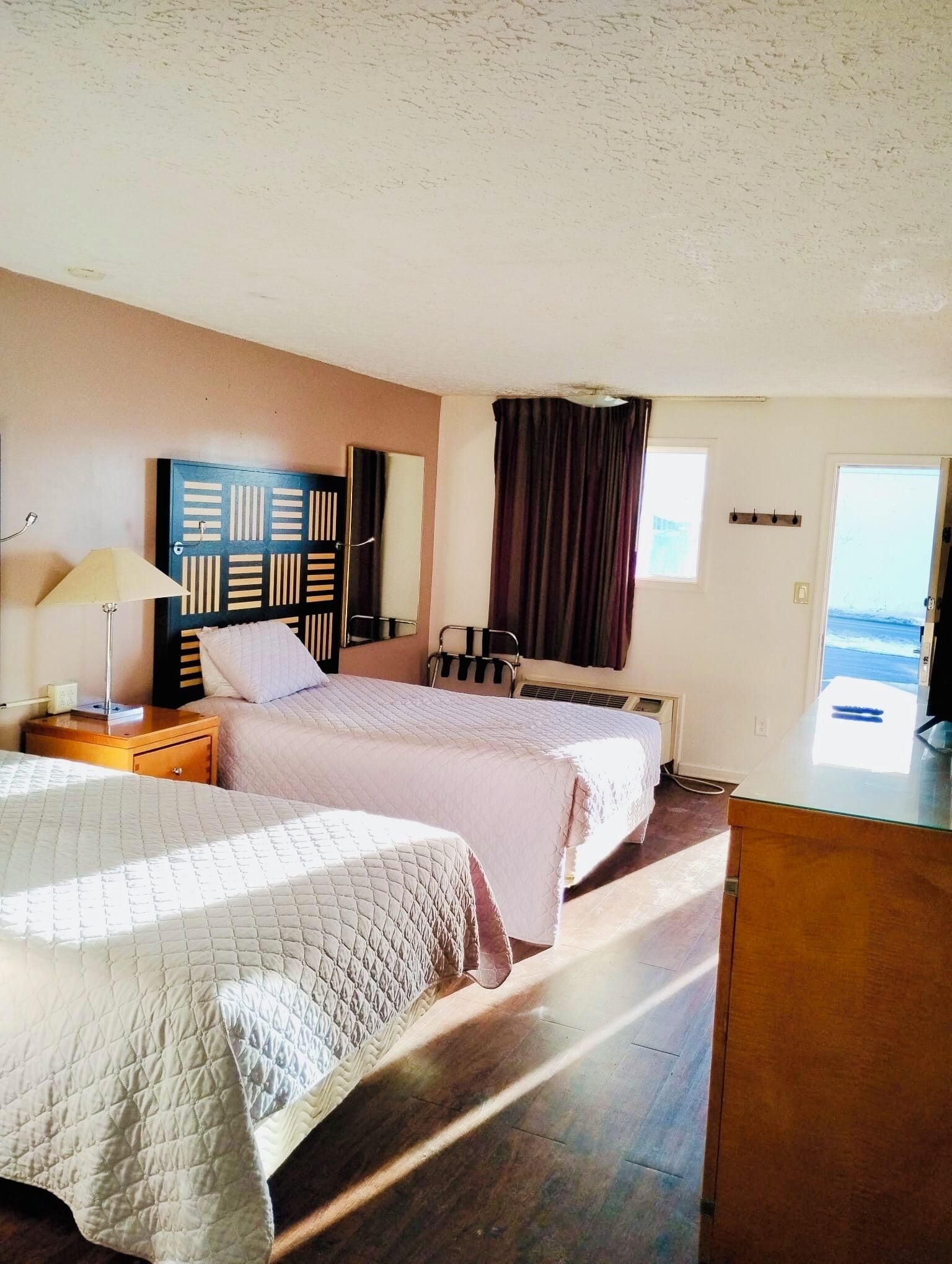 Quarto de hotel com duas camas, penteadeira de madeira, mesa de cabeceira com abajur, espelho e vista para o mar pela janela.