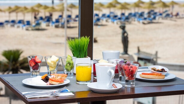 Mesa de café da manhã com diversas bebidas e alimentos, com vista para uma praia com guarda-sóis e cadeiras de praia.
