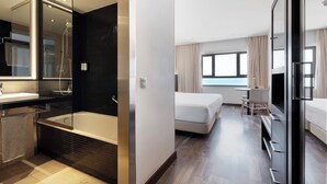 Quarto de hotel moderno com banheiro que inclui banheira, um espelho grande e chuveiro. O quarto tem uma cama, uma escrivaninha e uma televisão.
