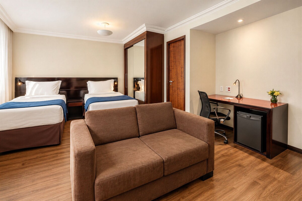 Quarto de hotel com duas camas, um sofá, uma escrivaninha e uma cadeira.