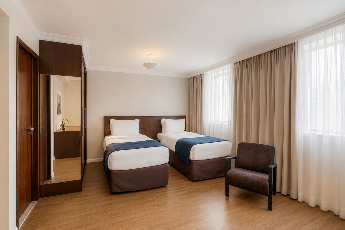 Quarto de hotel com duas camas, uma cadeira, um espelho e uma janela com cortinas.