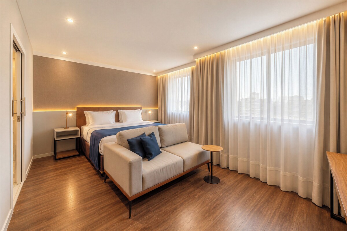 Quarto de hotel moderno com cama, sofá, mesa pequena e vista para a cidade através de amplas janelas.