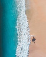 Vista aérea de uma praia com ondas quebrando na costa e uma pessoa segurando uma prancha de surf.