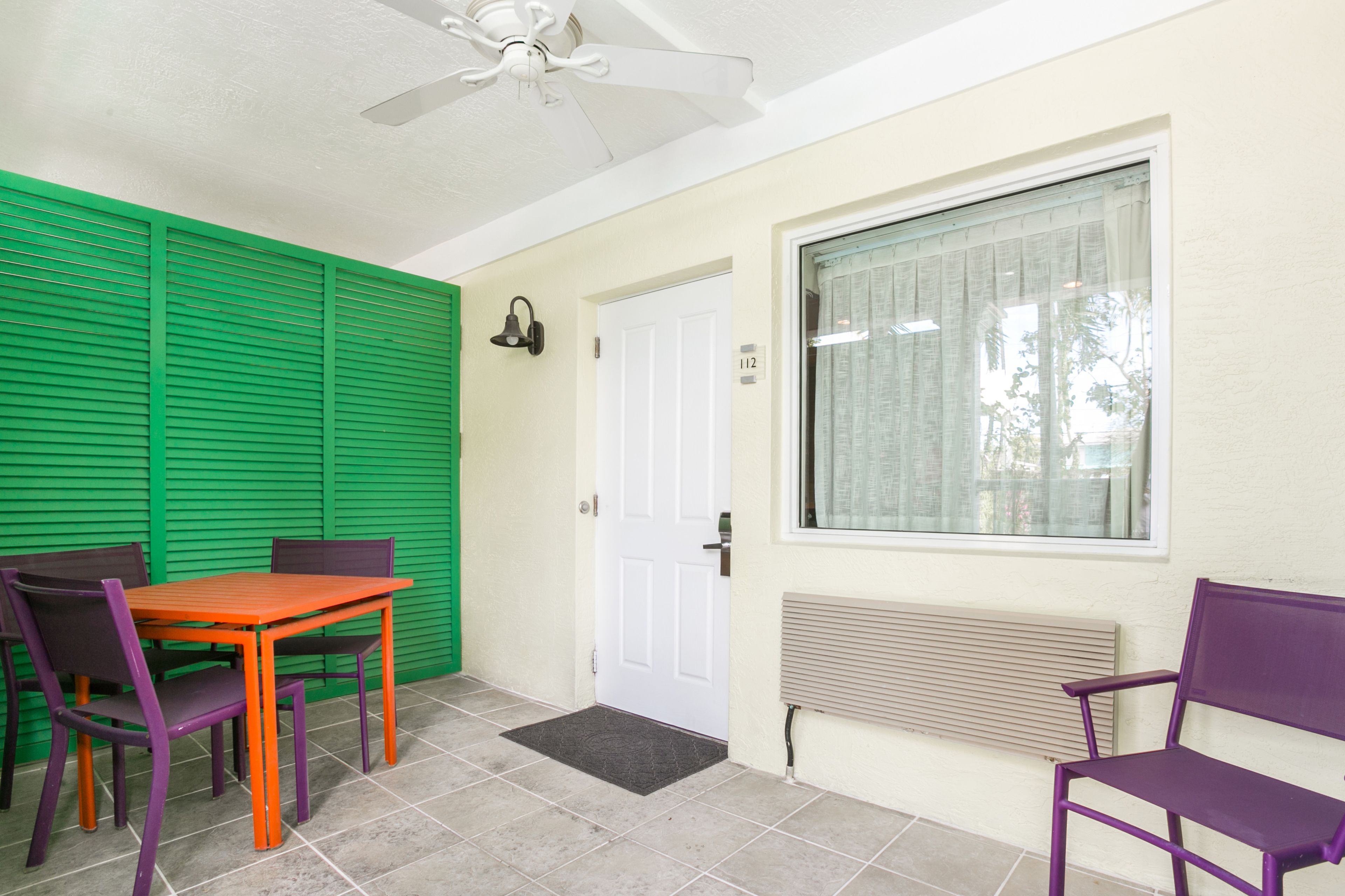 Um quarto com uma mesa e cadeiras, uma porta branca, uma janela com vista e um ventilador de teto.