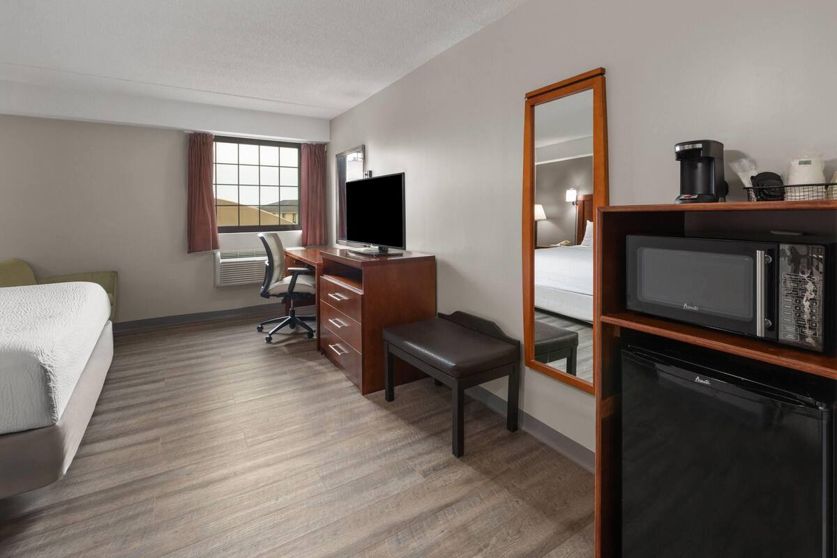 Quarto de hotel com cama, escrivaninha, cadeira, televisão, frigorífico e micro-ondas, além de uma janela com cortinas.