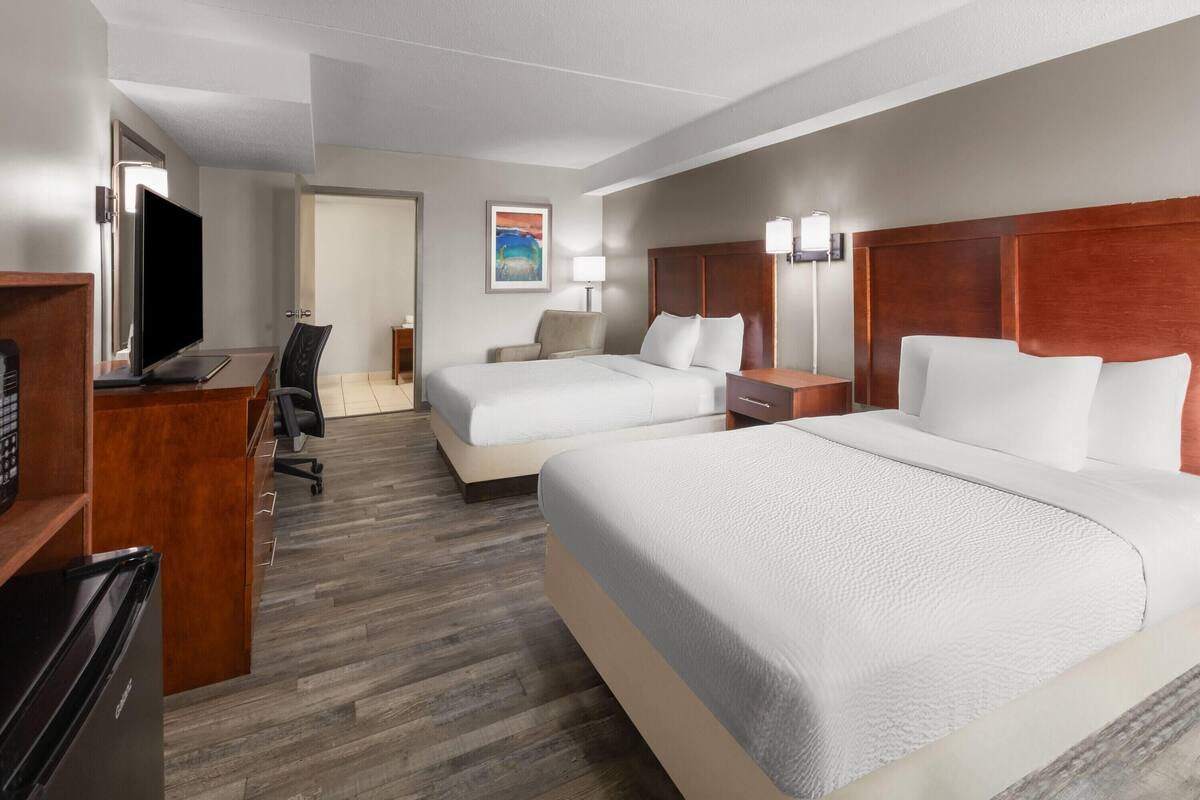 Quarto de hotel com duas camas, uma escrivaninha com computador, televisão e abajur.