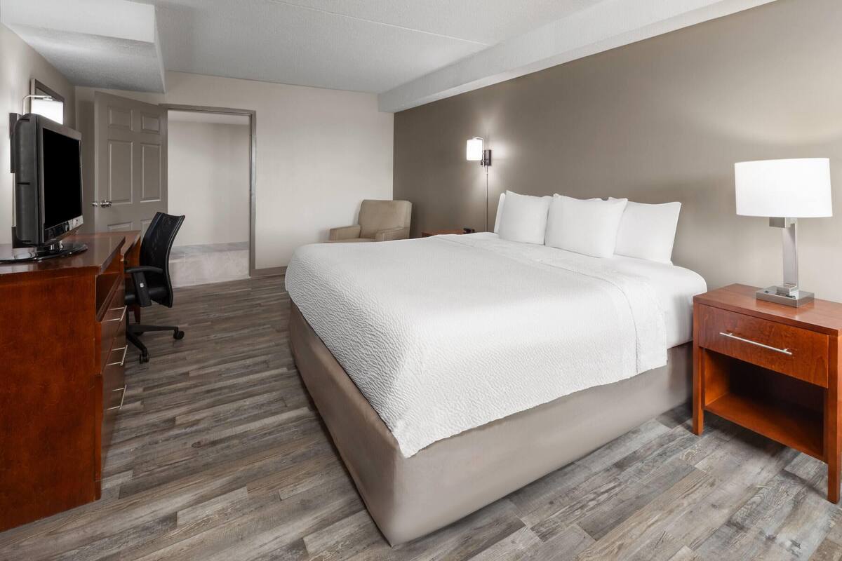 Quarto de hotel com uma cama grande, uma mesa com computador, uma cadeira e uma mesa de cabeceira com abajur.