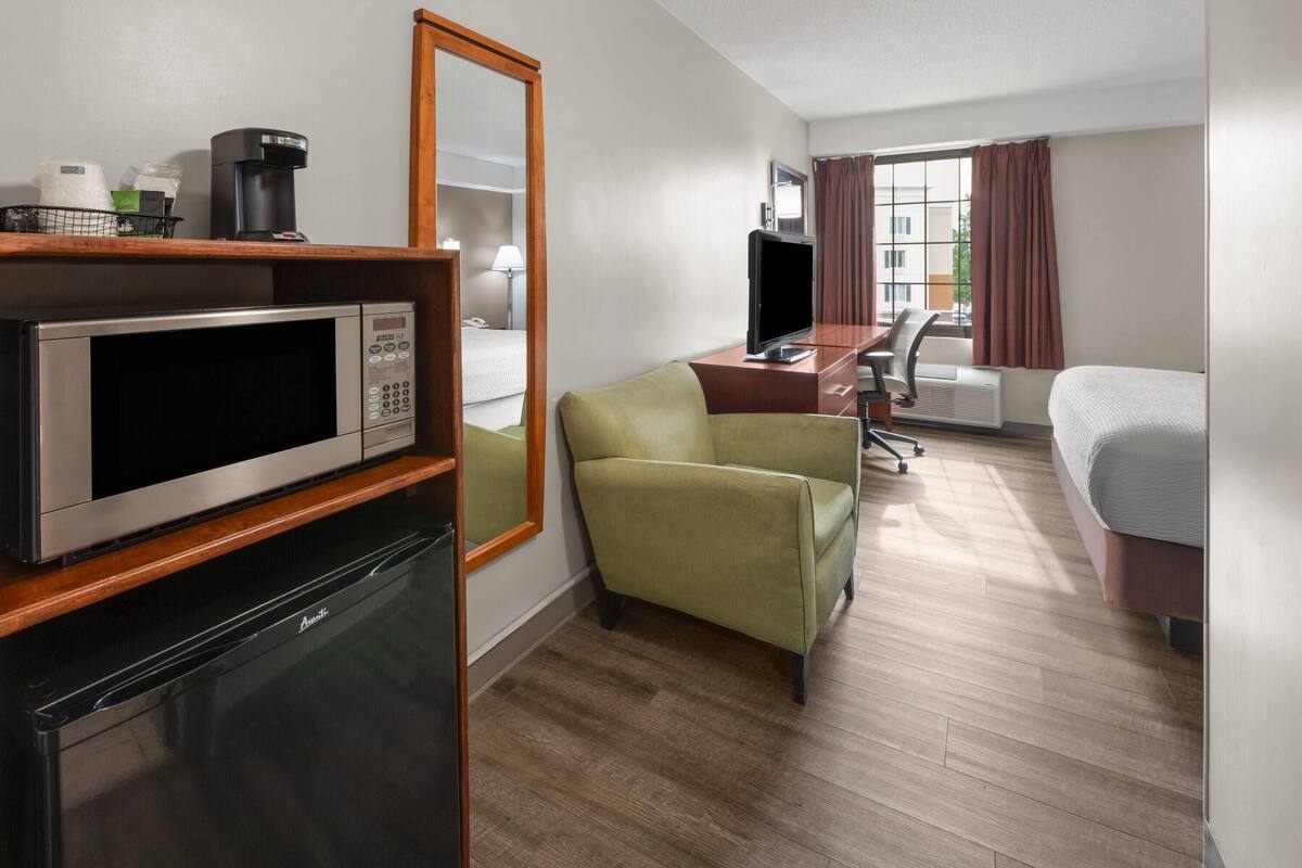 Quarto de hotel com micro-ondas, mini-frigorífico, poltrona verde, escrivaninha com computador, televisão e cama.