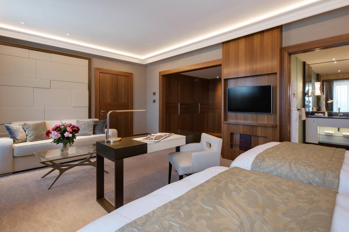 Quarto de hotel moderno com sofá, uma mesinha de centro com abajur, cama com roupa de cama estampada, TV fixada na parede e guarda-roupa de madeira.