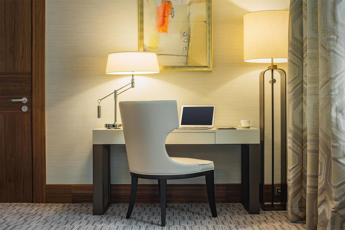 Quarto de hotel com escrivaninha, laptop, cadeira, duas luminárias, um quadro na parede e carpete estampado.
