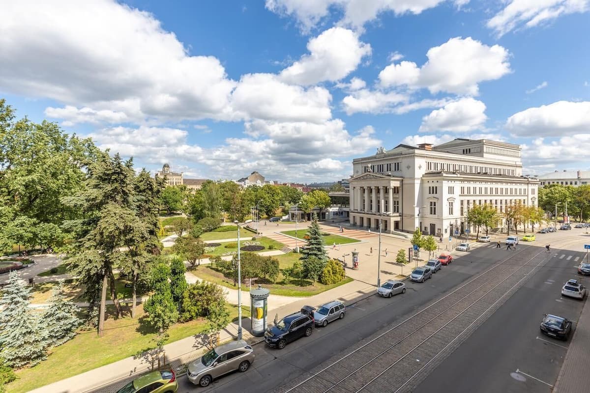 Um edifício grande e histórico com arquitetura clássica, cercado por um parque bem cuidado e uma linha de bonde.