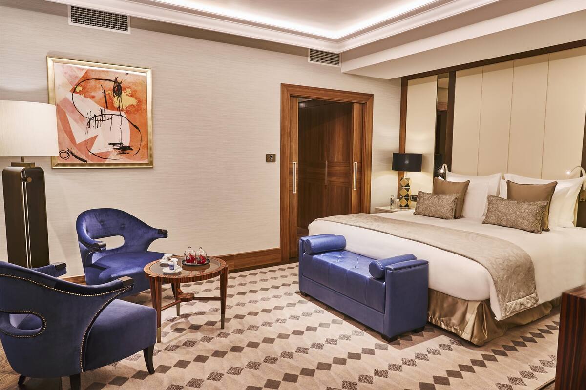Quarto de hotel com uma cama grande, um sofá azul, uma mesinha de centro com uma luminária e um quadro na parede.
