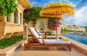 Uma área à beira da piscina com duas espreguiçadeiras, uma mesa com guarda-sol amarelo, e um muro decorativo com plantas e flores.