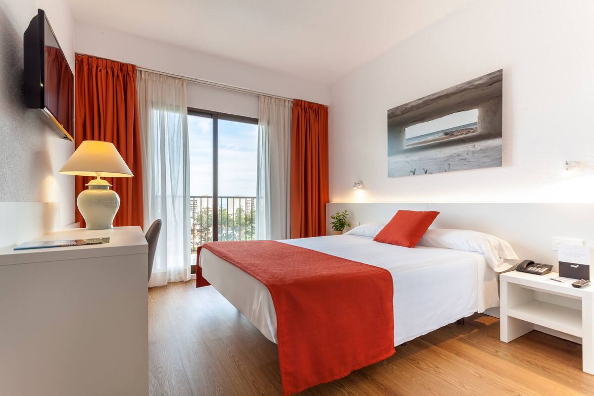 Quarto de hotel com cama, escrivaninha, cadeira, mesa de cabeceira, luminária e um quadro na parede.