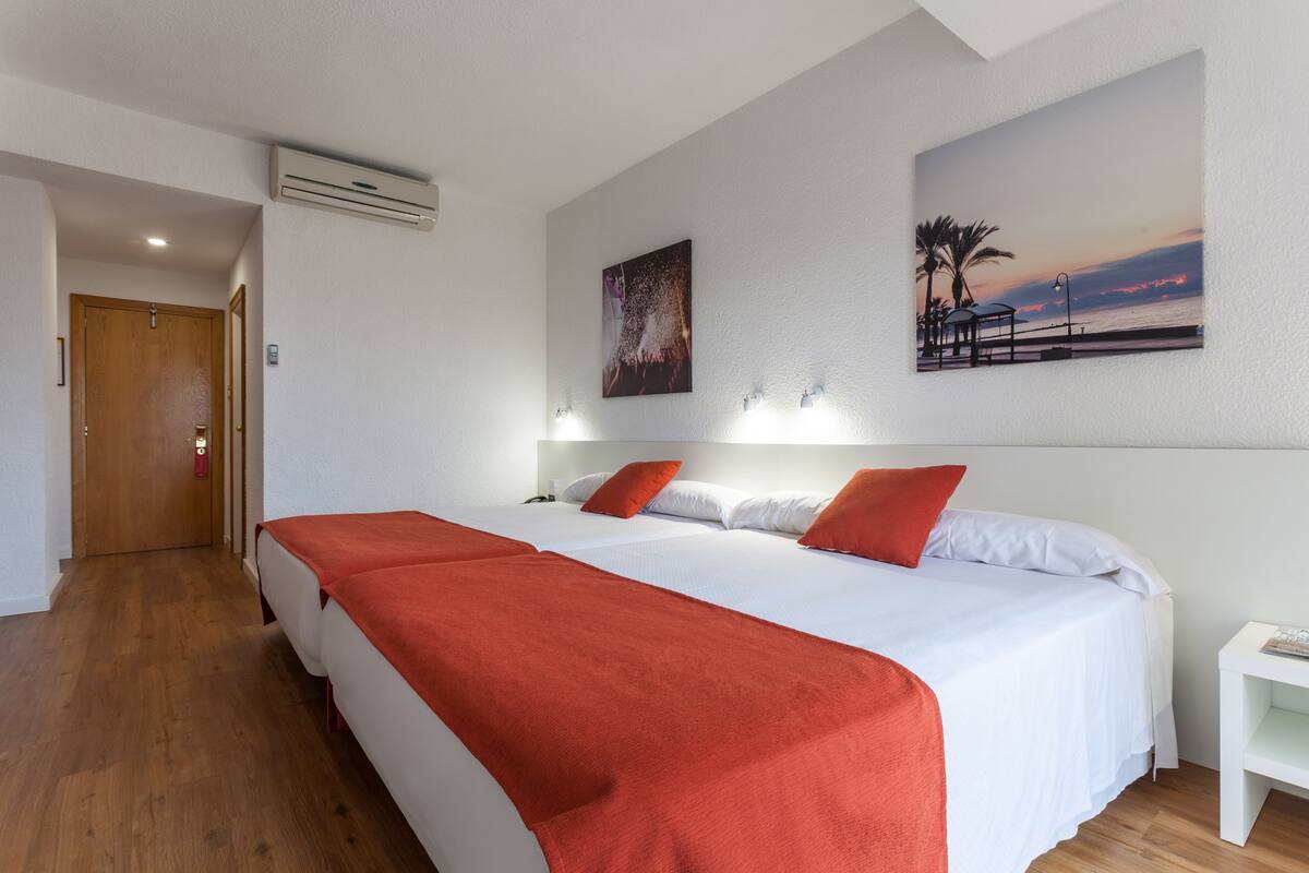 Quarto de hotel com duas camas, um quadro de um pôr do sol e um ar condicionado.