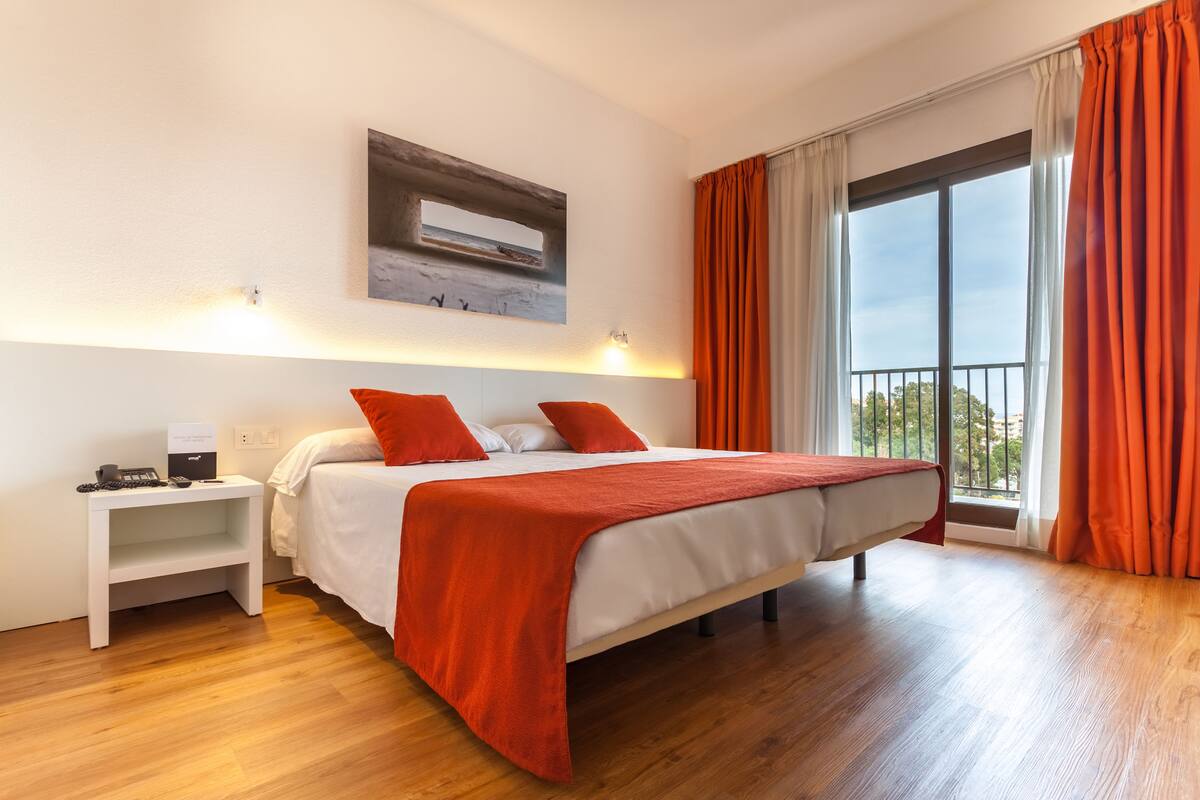 Quarto de hotel moderno com uma cama grande, uma mesa de cabeceira e uma janela com cortinas.