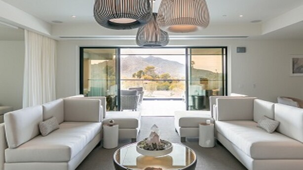 Uma sala de estar moderna com uma mesa de centro de vidro, dois sofás brancos e vista para montanhas através de grandes portas de vidro.