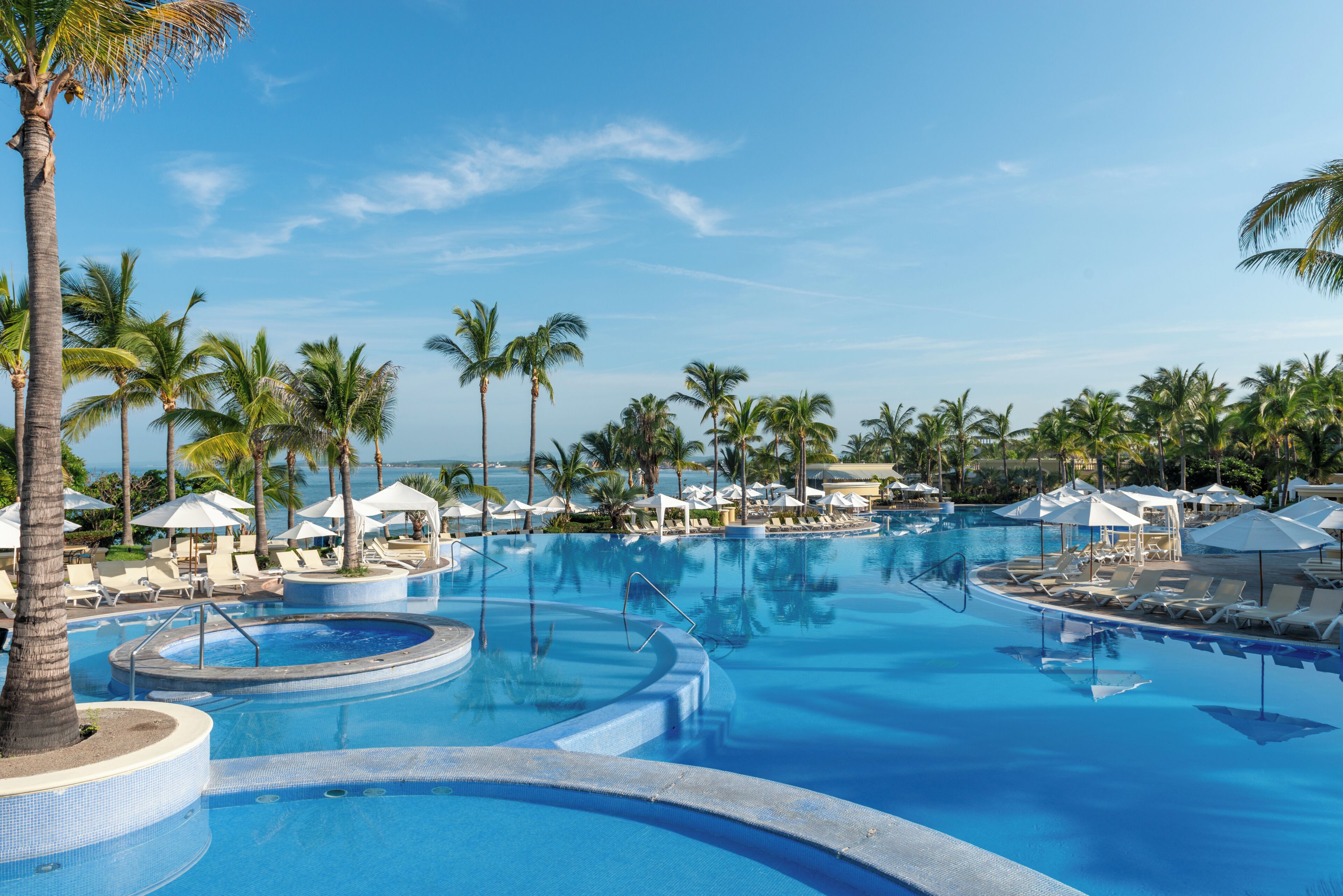 Uma grande piscina ao ar livre com vários níveis e palmeiras ao redor.
