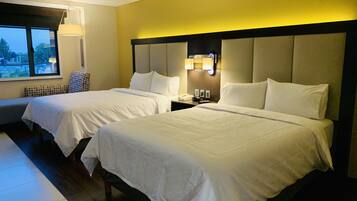 Quarto de hotel com duas camas, mesa de cabeceira com abajur, janela com vista e um pequeno espaço de estar.