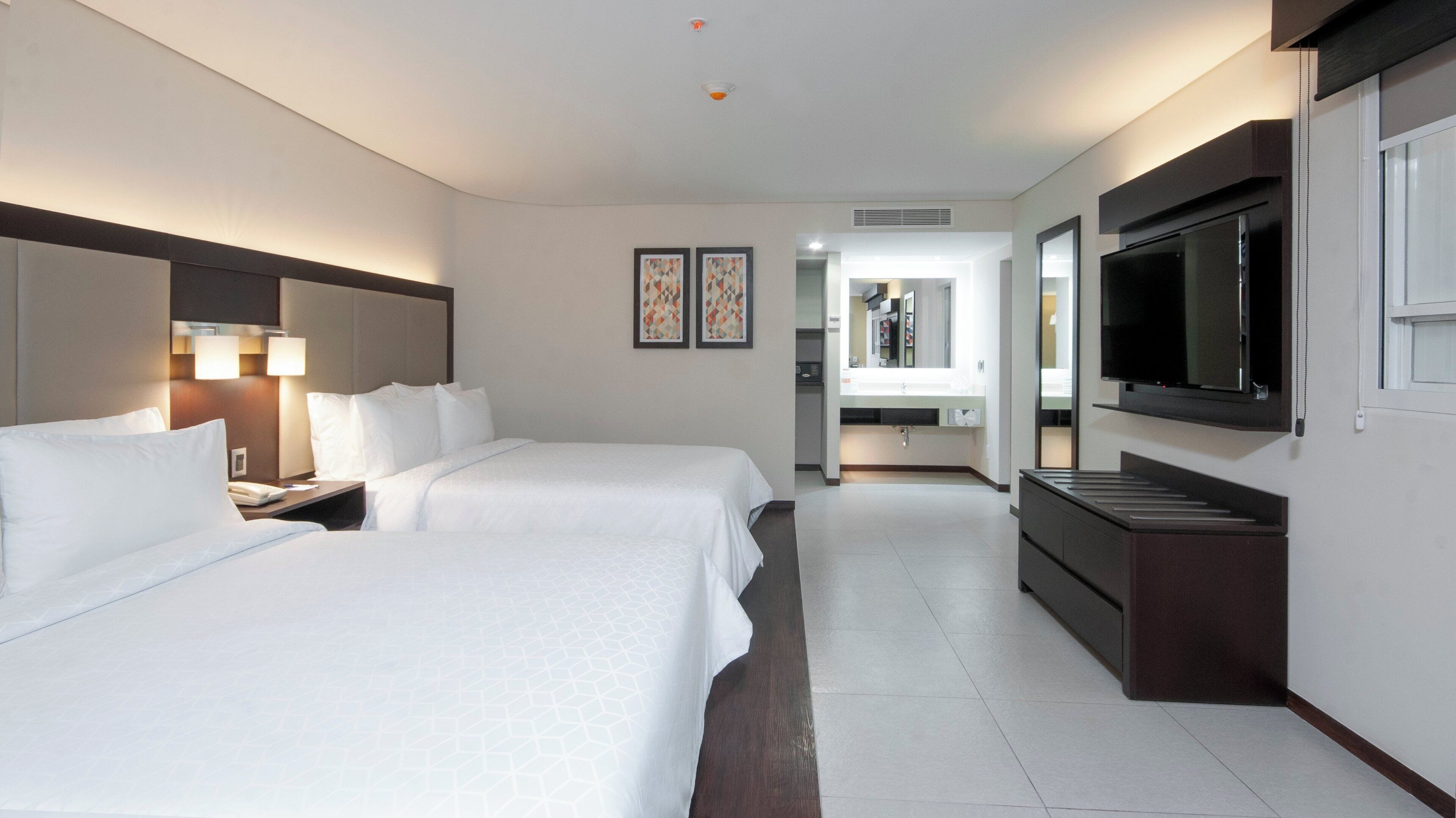 Quarto de hotel com duas camas, televisão, escrivaninha e banheiro visível por uma porta aberta.