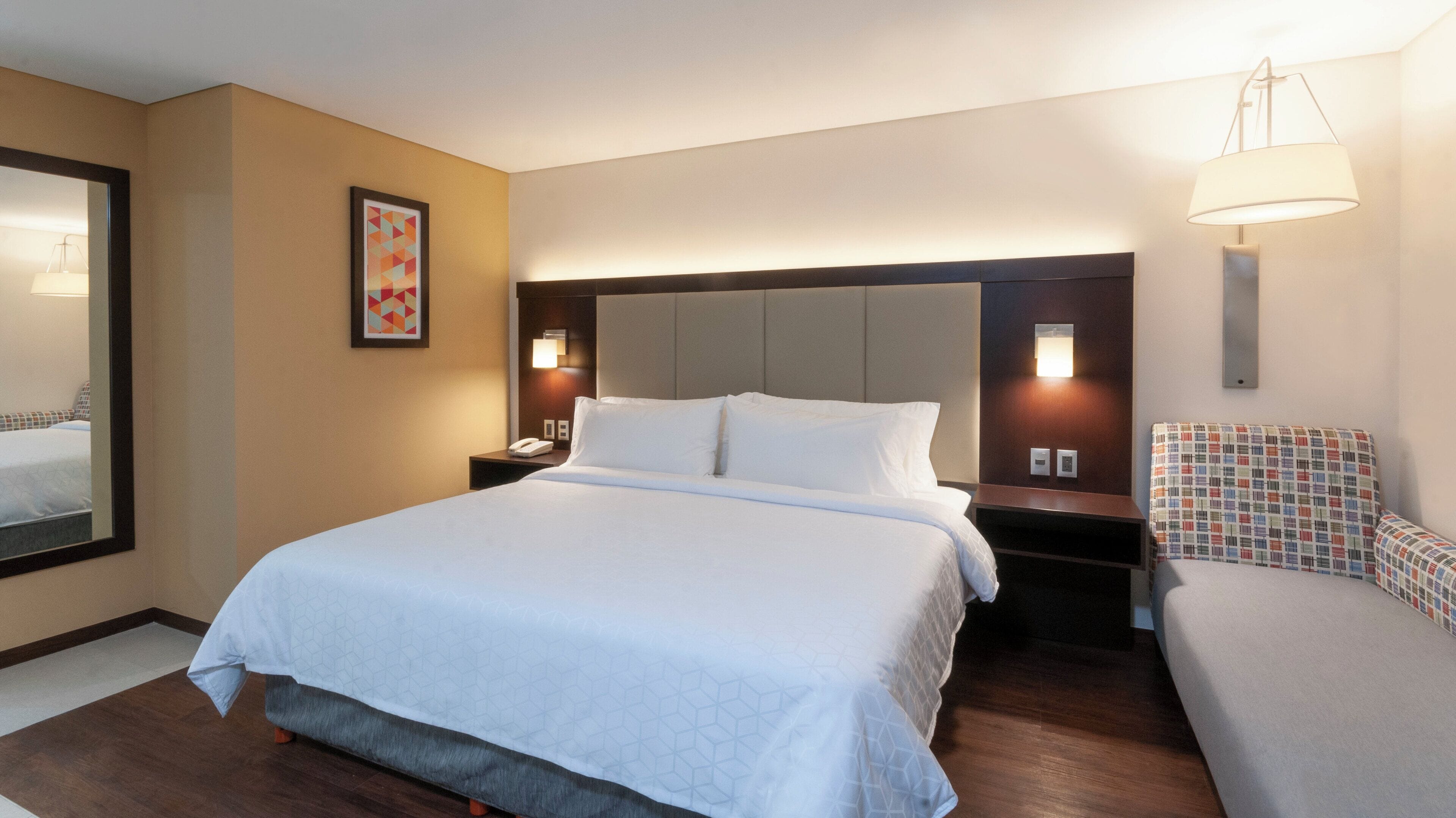 Quarto de hotel com cama grande, mesas de cabeceira, abajur, espelho e luz na parede.