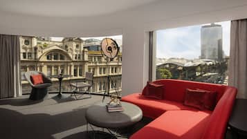 Quarto de hotel moderno com um sofá vermelho, uma poltrona cinzenta e uma pequena mesa redonda. Uma grande janela oferece vista para um edifício histórico e um arranha-céu imponente.
