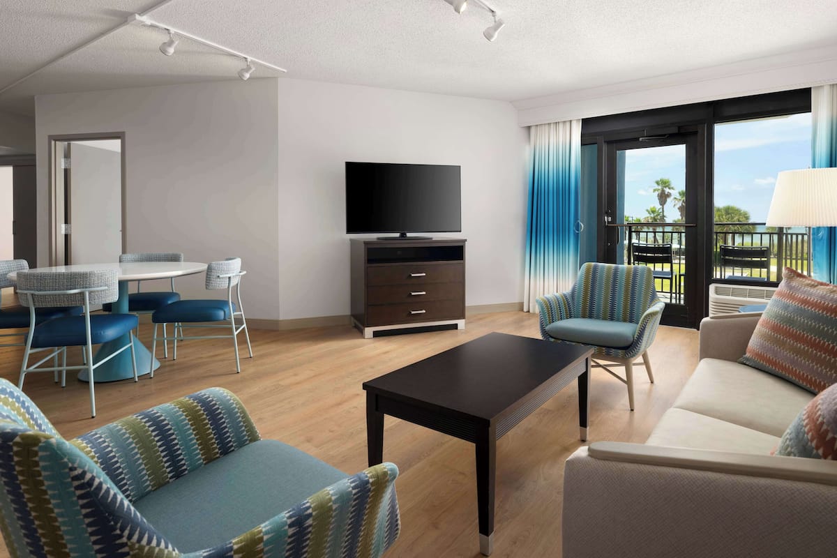 Quarto de hotel moderno com televisão de tela plana, área de refeições com mesa redonda e cadeiras, sofá e varanda com vista.