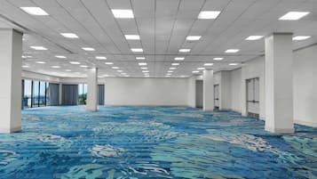 Uma sala de conferências grande e vazia, com carpete azul estampado, paredes brancas e uma grade de luminárias no teto.