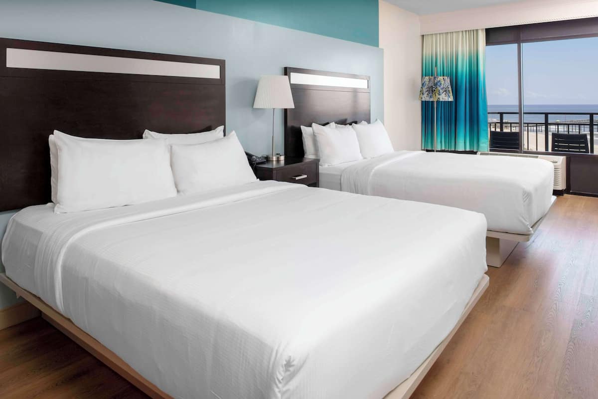 Quarto de hotel com duas camas, uma mesinha de cabeceira, uma luminária e vista para o oceano.