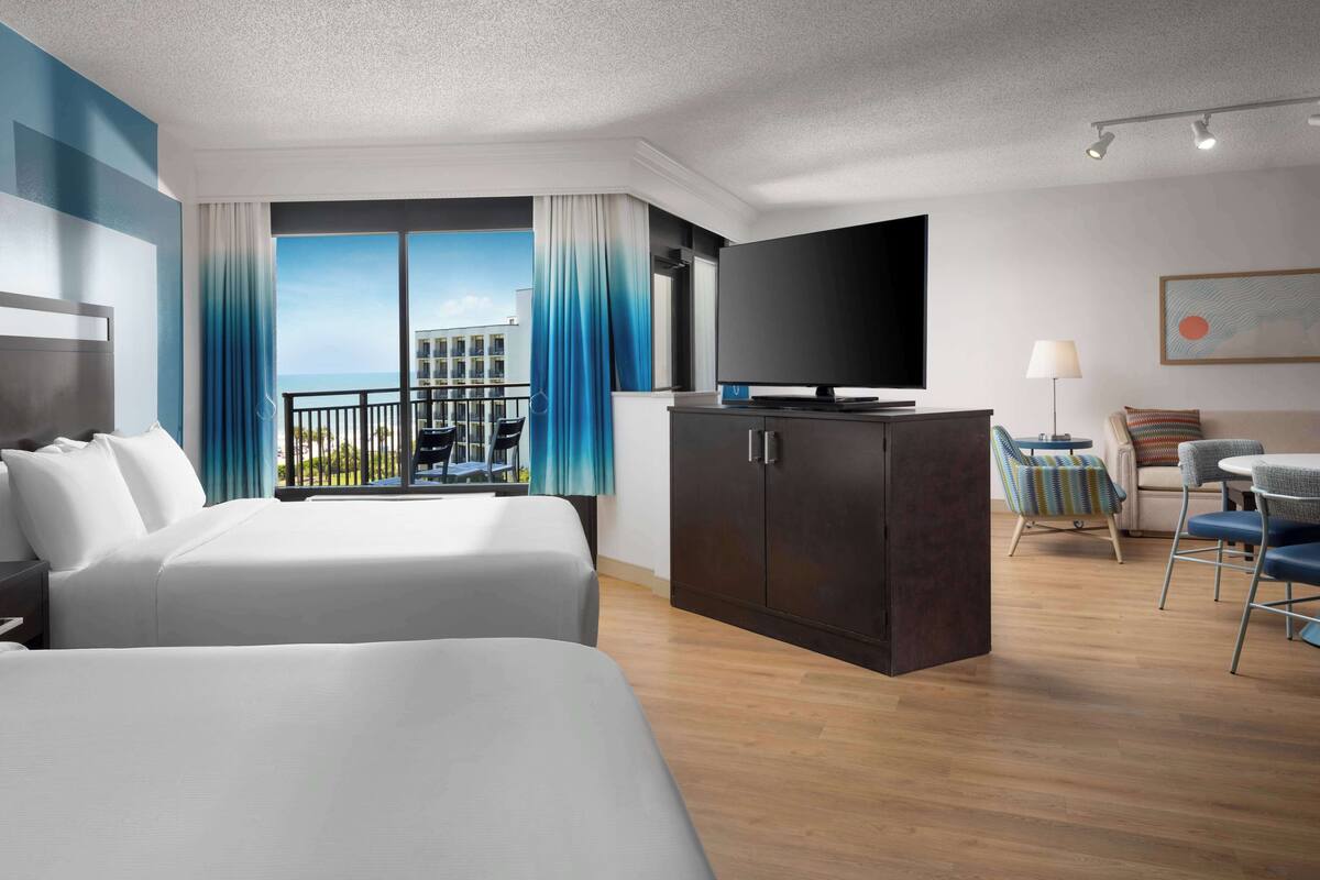 Quarto de hotel moderno com duas camas, televisão, varanda com vista e área de jantar com cadeiras e mesa.