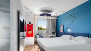 Quarto de hotel moderno com uma cama, uma escrivaninha, uma cadeira, uma televisão, uma janela com cortinas e uma parede azul.