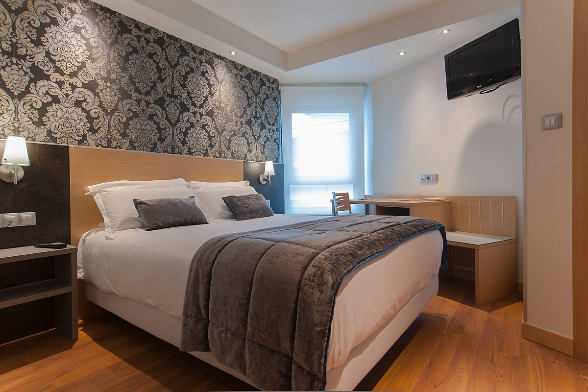 Quarto de hotel moderno com uma cama grande, mesas de cabeceira, uma escrivaninha e uma TV fixada na parede.