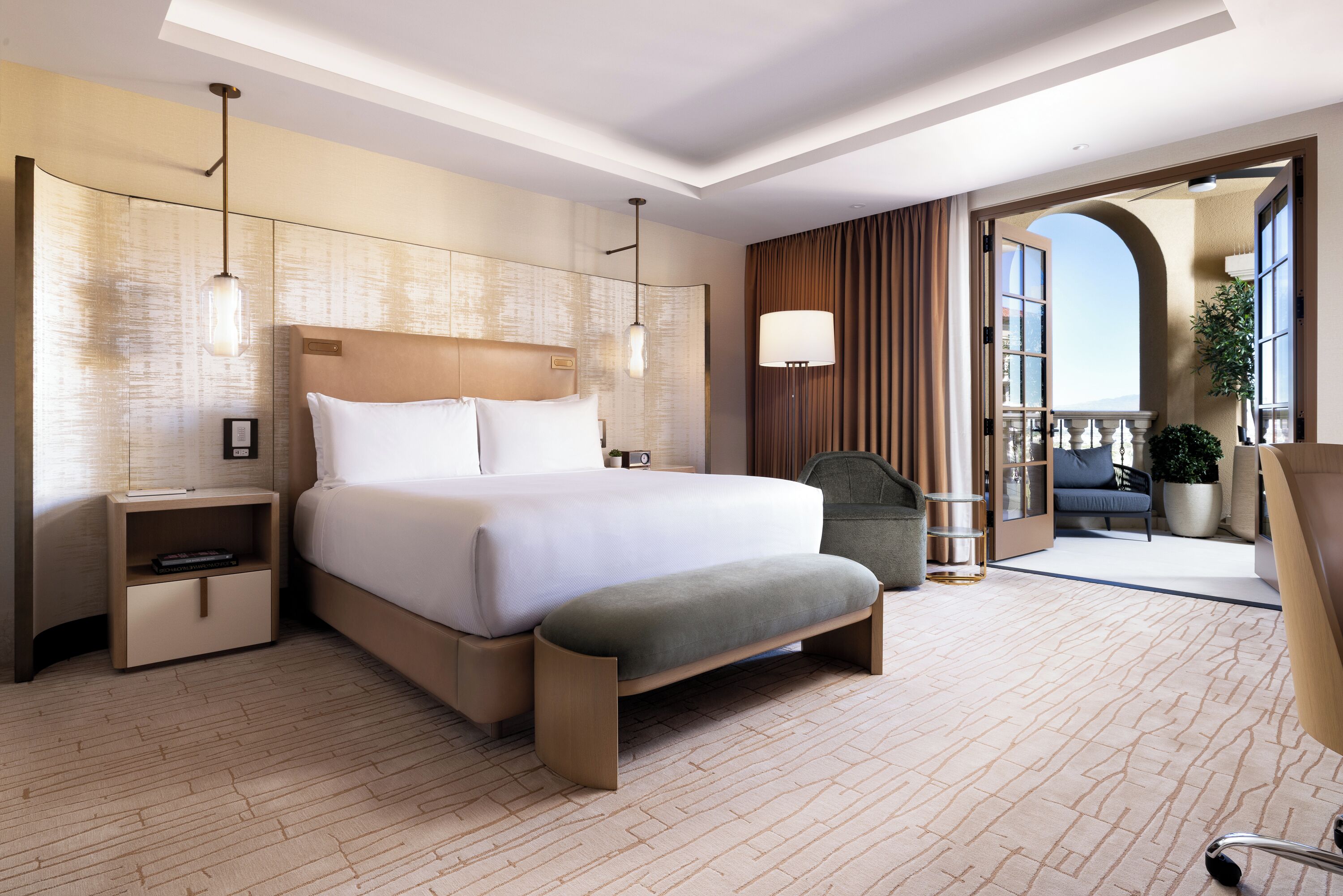 Quarto de hotel com cama, mesas de cabeceira, um banco, uma cadeira, um abajur e vista através de uma porta aberta.