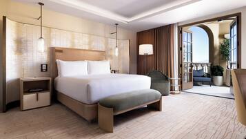 Quarto de hotel com cama, mesas de cabeceira, um banco, uma cadeira, um abajur e vista através de uma porta aberta.