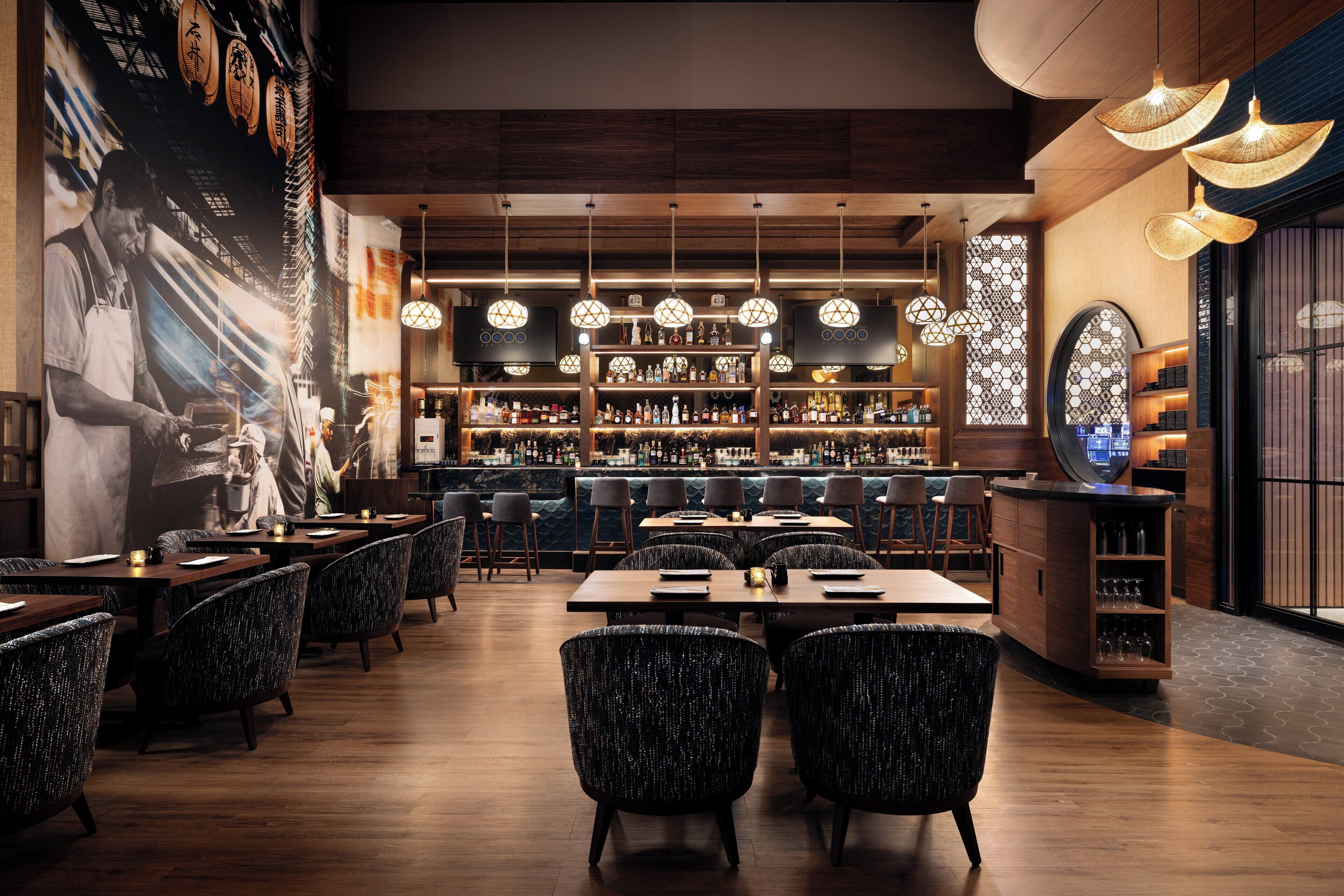 Um restaurante com mesas e cadeiras de madeira, área de bar com prateleiras cheias de garrafas e luminárias pendentes.