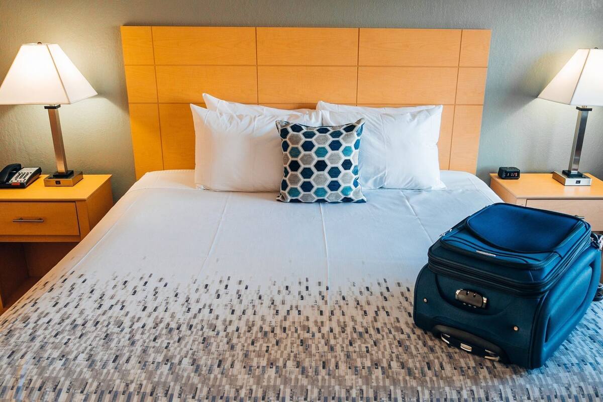 Quarto de hotel com cama, duas luminárias de cabeceira, relógio, telefone e mala.