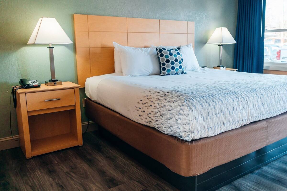 Quarto de hotel com uma cama grande, duas mesas de cabeceira com abajures, um painel de cabeceira de madeira e vista para a rua pela janela.