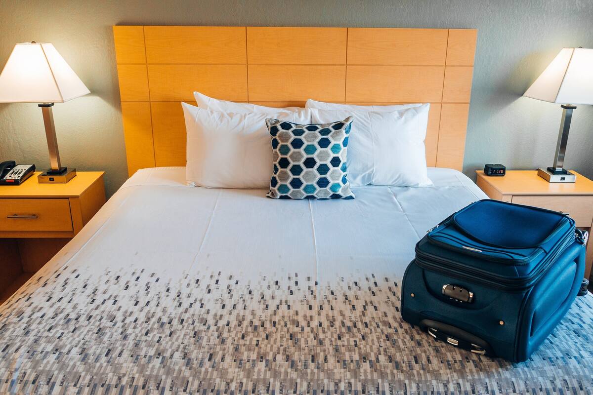 Quarto de hotel com uma cama, duas mesas de cabeceira com luminárias, um telefone, um relógio digital e uma mala azul sobre a cama.