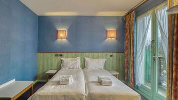 Uma cama de casal com duas almofadas e um cobertor, uma mesinha de cabeceira, uma pequena mesa e uma janela com cortinas.