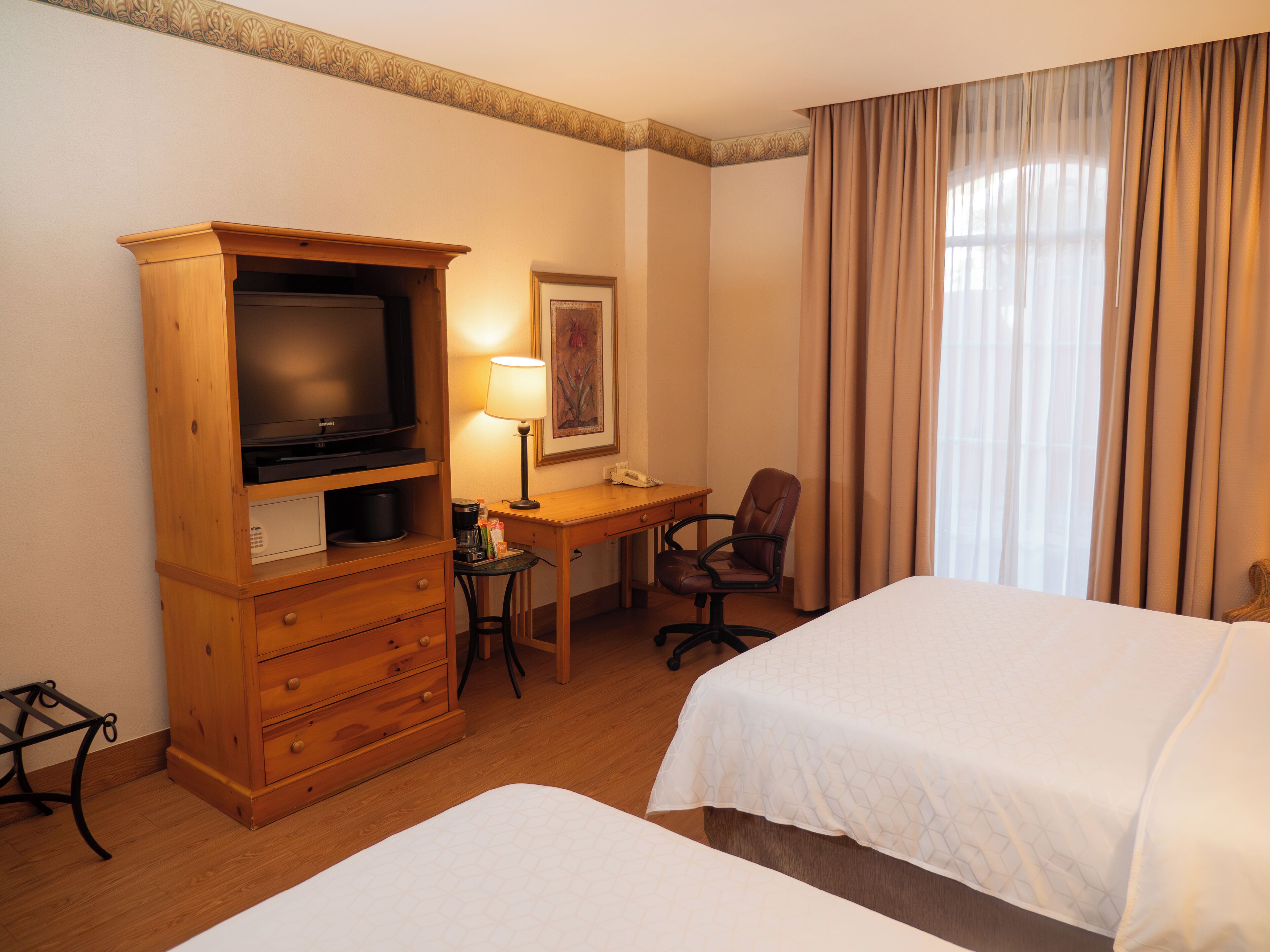 Quarto de hotel com duas camas, um suporte de TV de madeira, uma escrivaninha com cadeira e uma janela grande com cortinas.