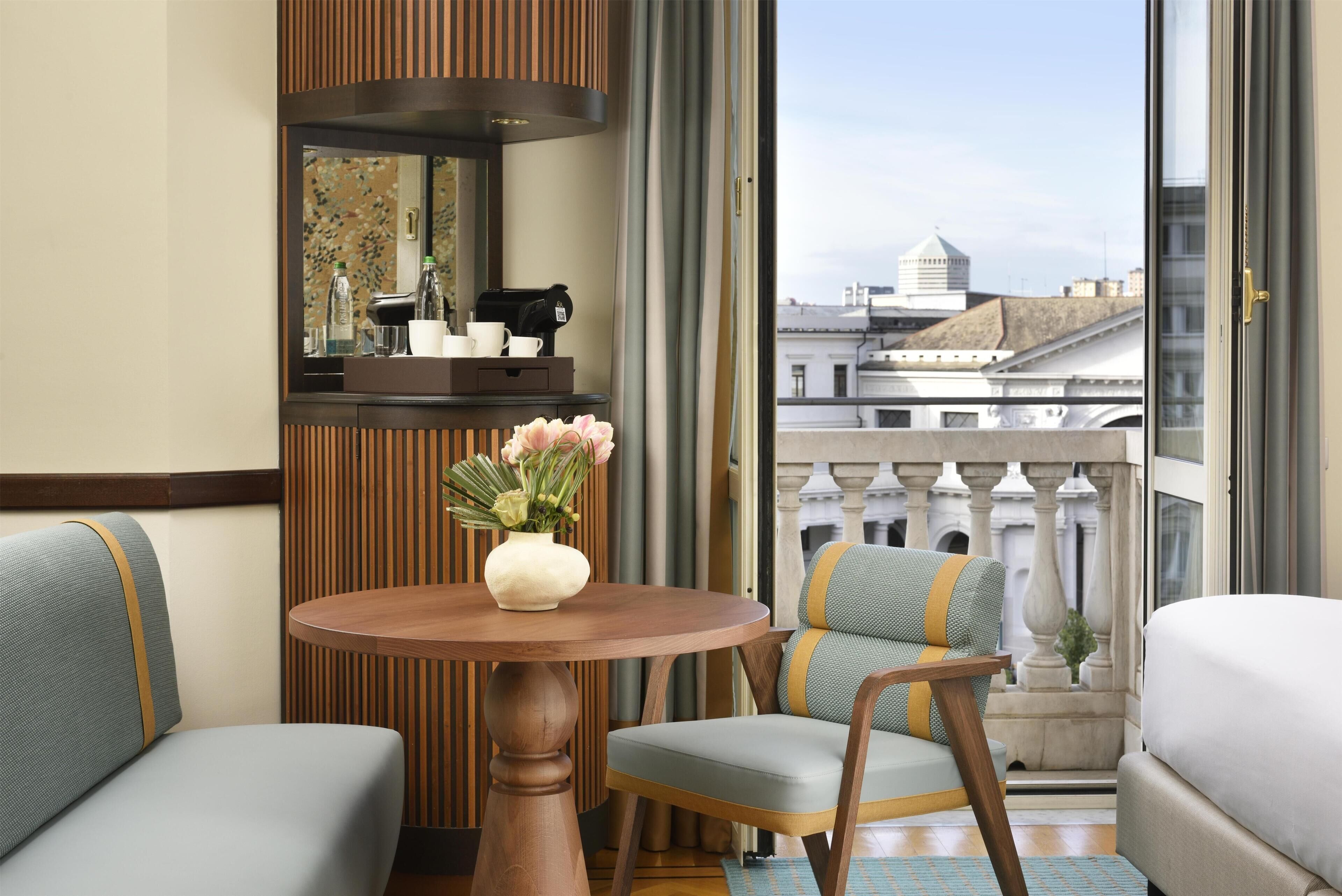 Quarto de hotel com varanda, uma mesa redonda de madeira com um vaso, duas cadeiras estofadas e vista para um edifício com cúpula.