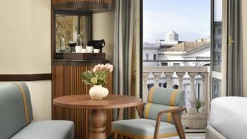 Quarto de hotel com varanda, uma mesa redonda de madeira com um vaso, duas cadeiras estofadas e vista para um edifício com cúpula.