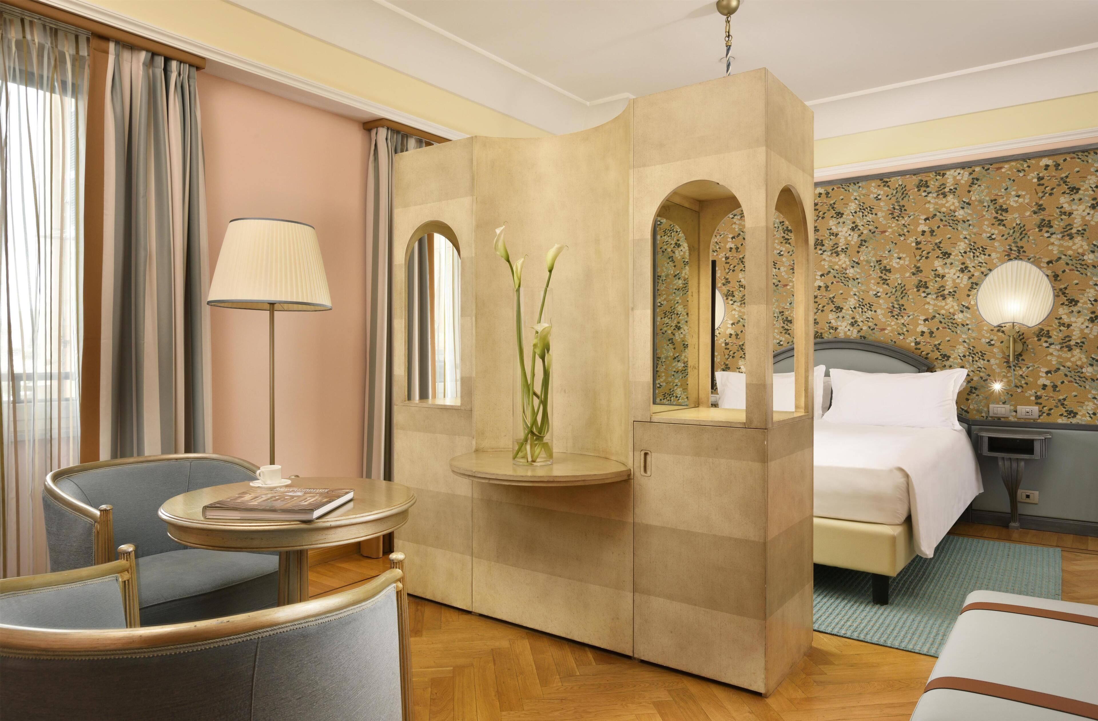 Quarto de hotel com cama, mesa redonda com um livro, cadeira, abajur e um vaso com flores.
